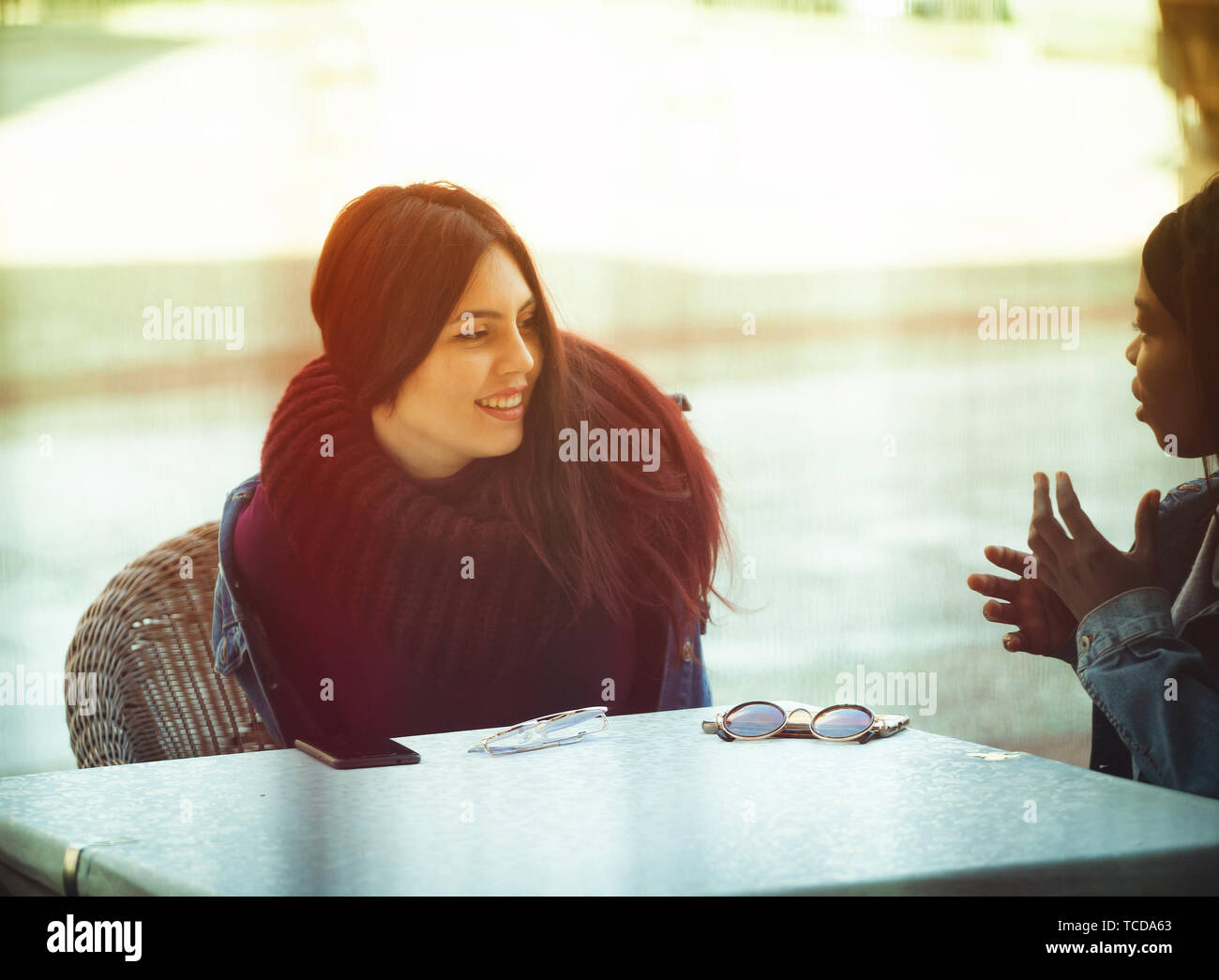 Two young beautiful girls sit at a table talking and using the mobile ...