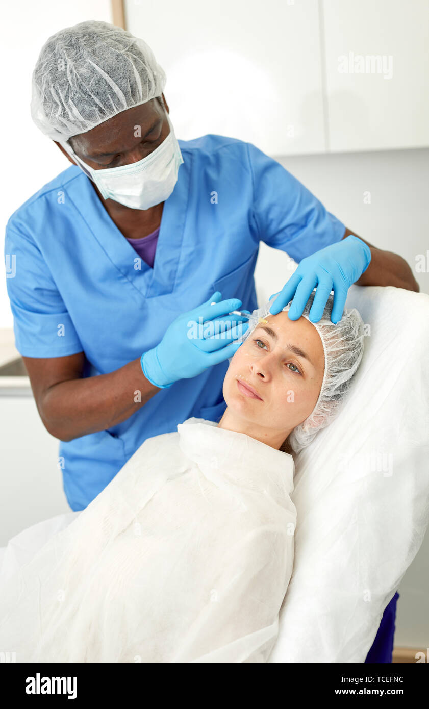 Male doctor in mask is doing injection to woman patient in skin of face ...