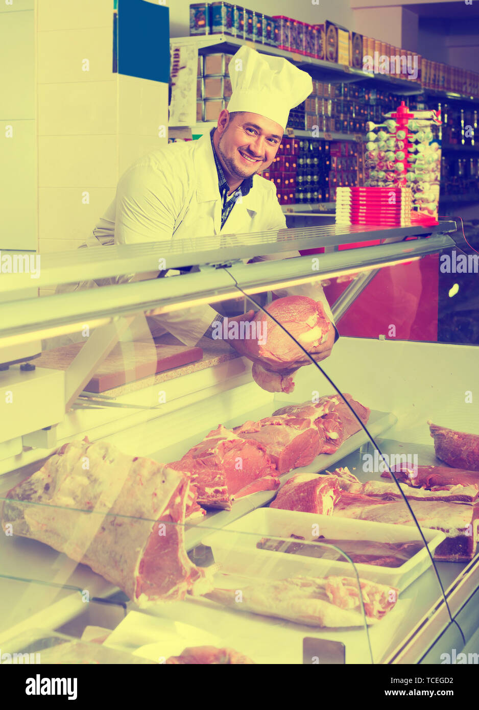 Professional smiling young butcher cutting fresh lamb meat in workshop ...