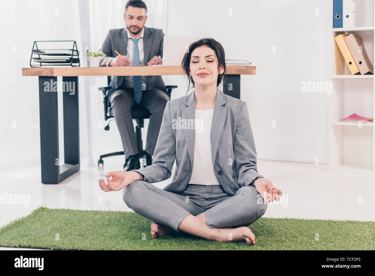 businesswoman meditating in Lotus Pose on grass mat while businessman ...