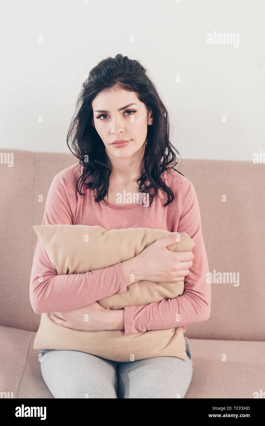 depressed woman sitting on couch, looking at camera and hugging pillow ...