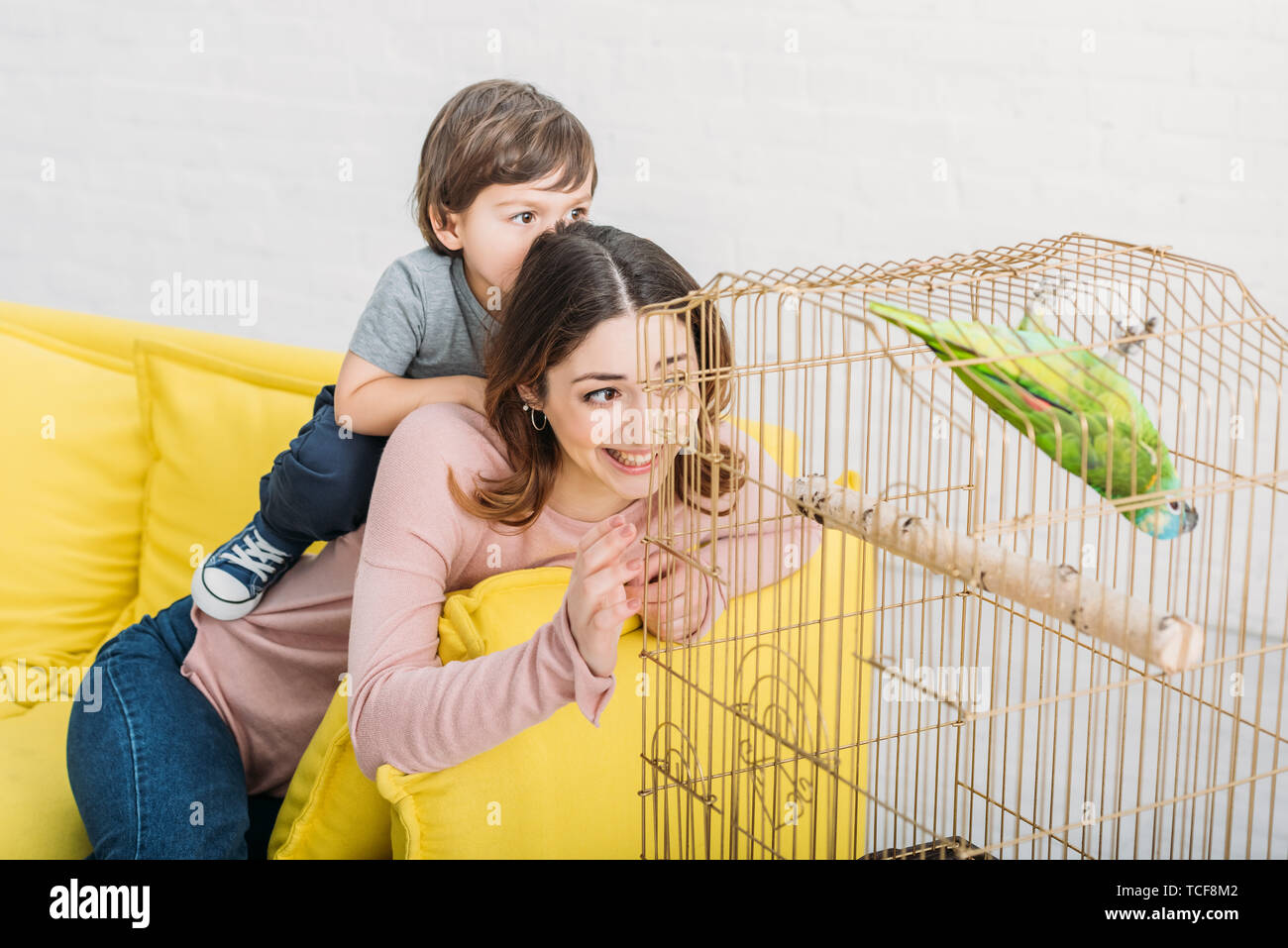 smiling mother and son looking at green parrot in bird cage at home ...