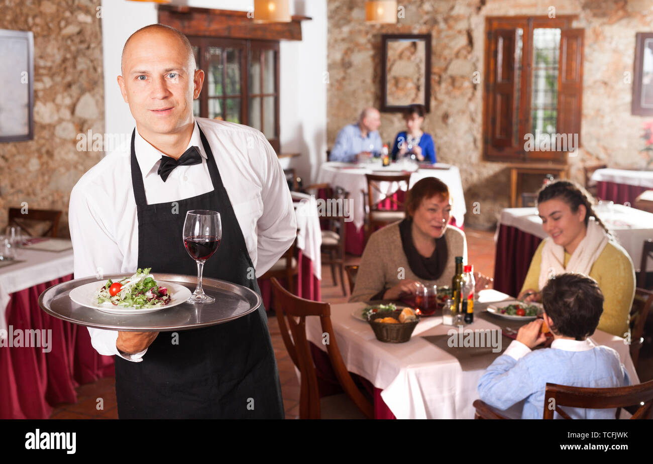 Positive waiter with tray in foreground and visitors having dinner at ...