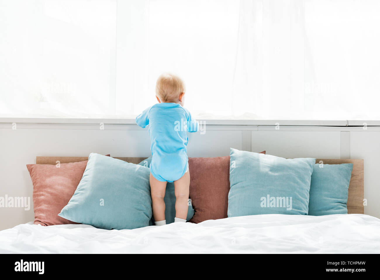 back view of toddler kid standing on bed and looking at window in ...