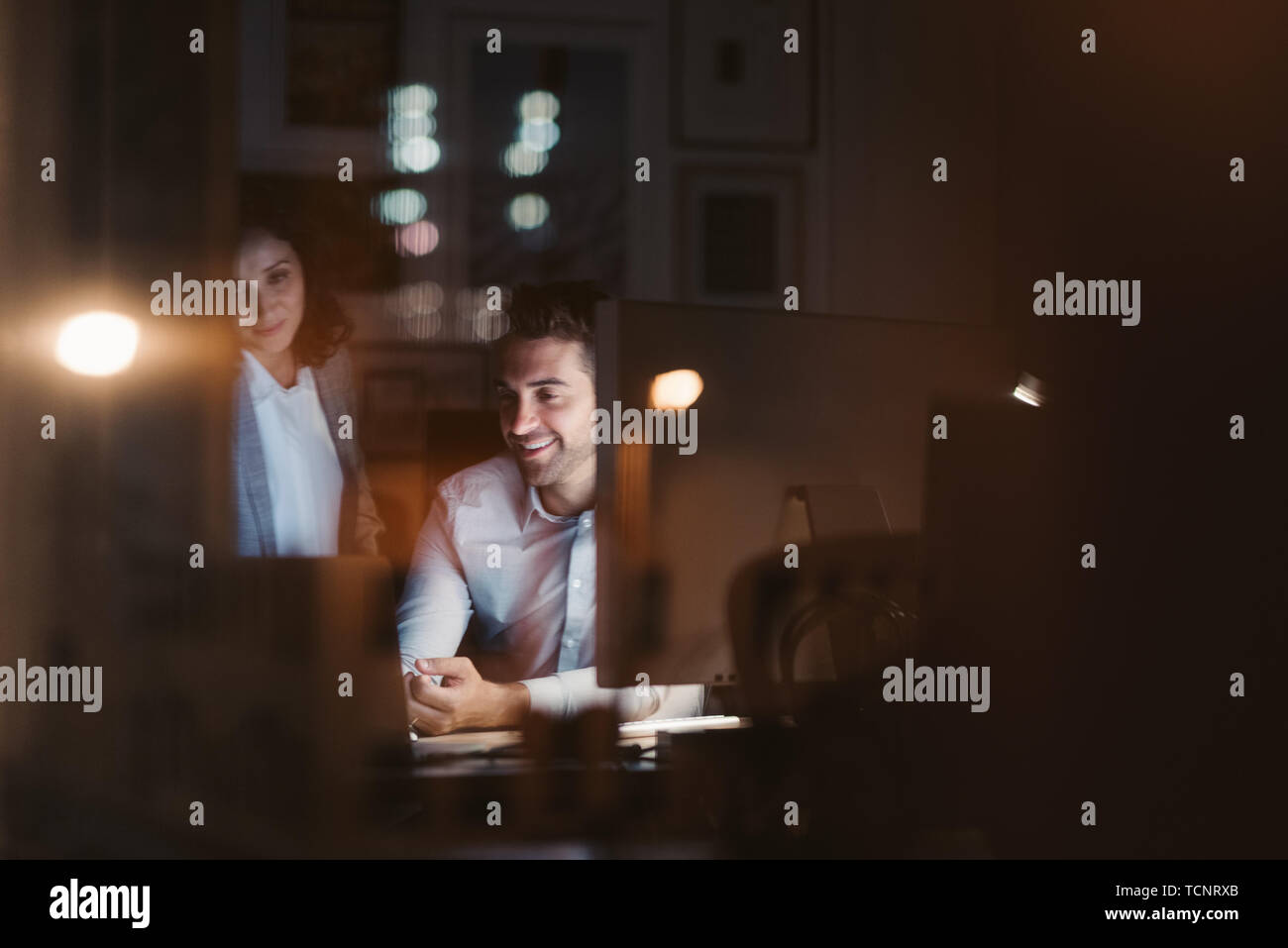 Two smiling young businesspeople working overtime together at a desk ...