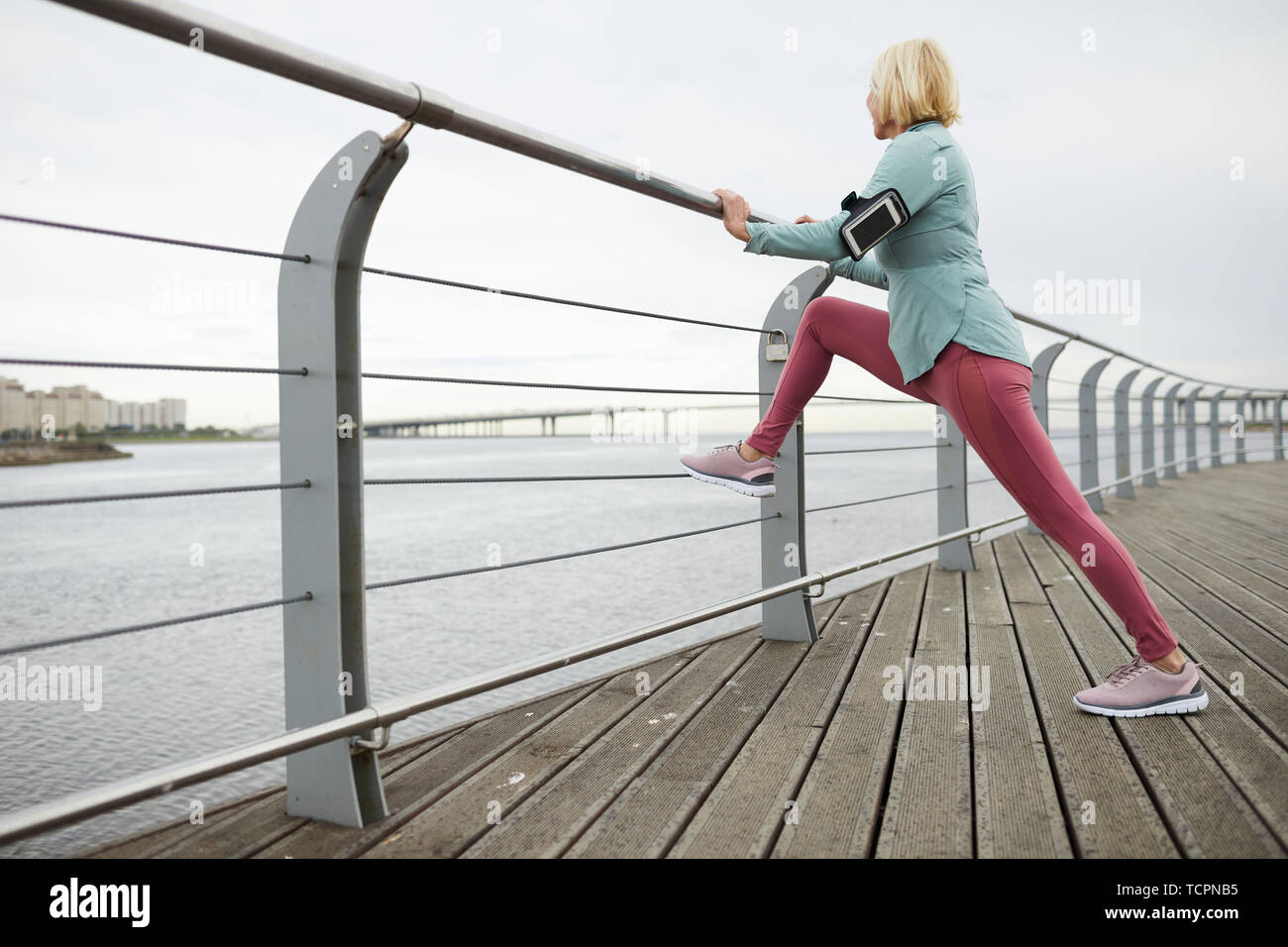 Blonde active woman in sportswear standing by banister and looking at river while doing ...