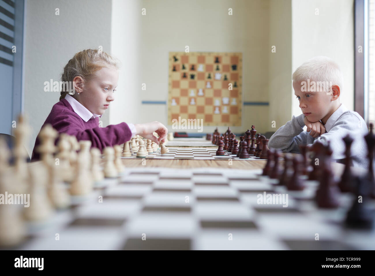 Blonde boy in white shirt and girl in purple sweater playing chess and ...