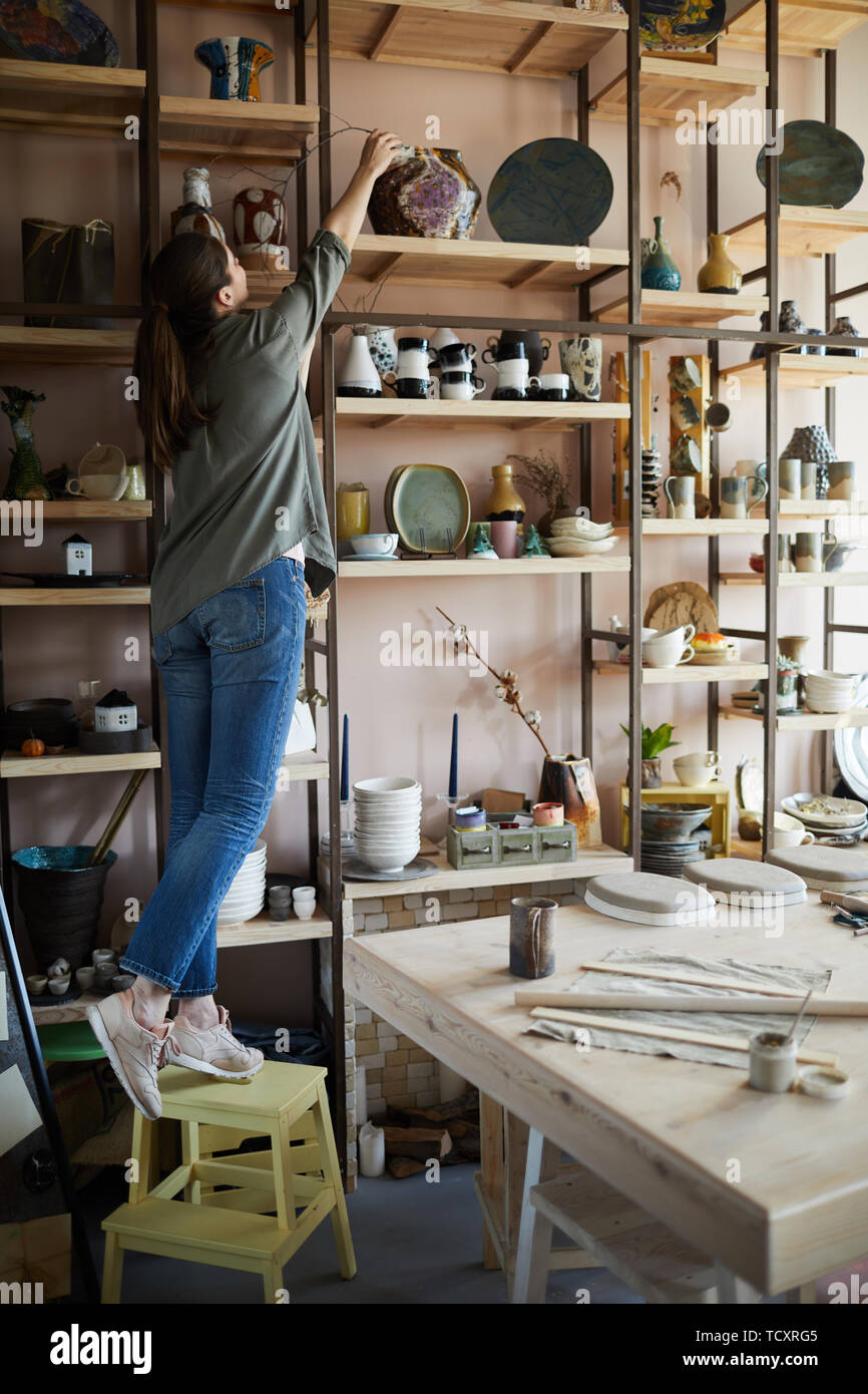 Full length portrait of young woman standing on stool reaching for ...