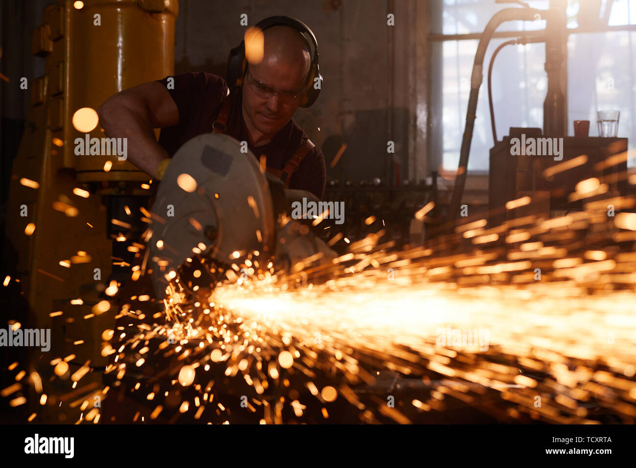 Serious skilled male blacksmith in ear protectors and safety goggles ...