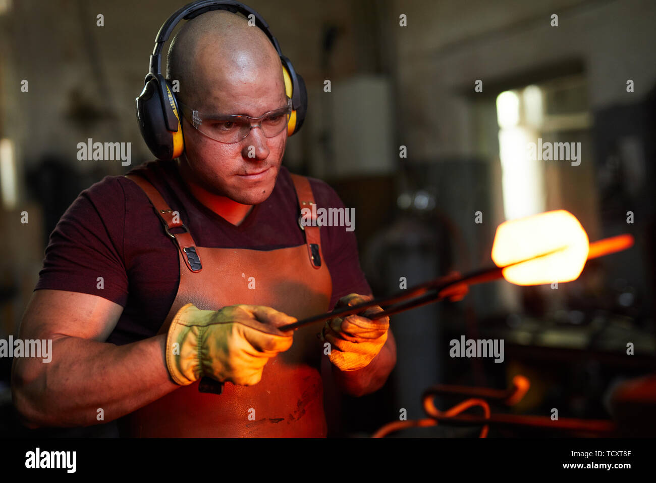 Serious bald young blacksmith in ear protectors and safety goggles ...