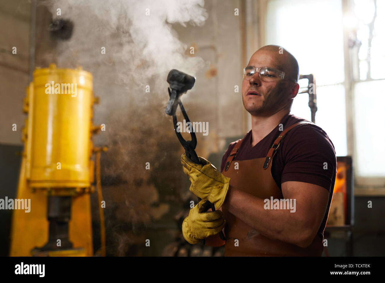 Pensive bald young blacksmith in safety goggles keeping metal part in ...