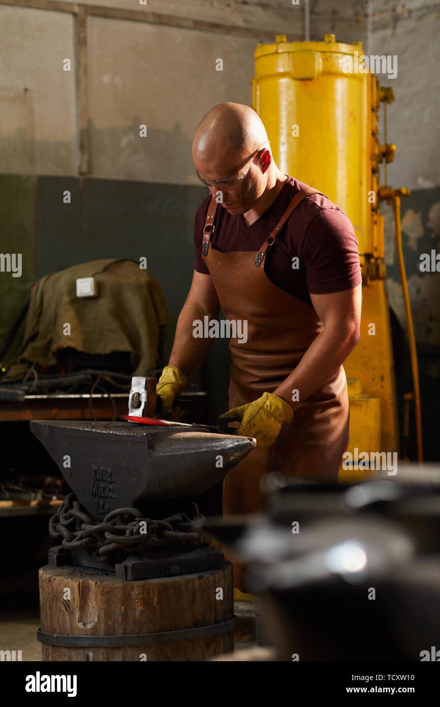Concentrated bald craft worker in apron standing at anvil and changing ...