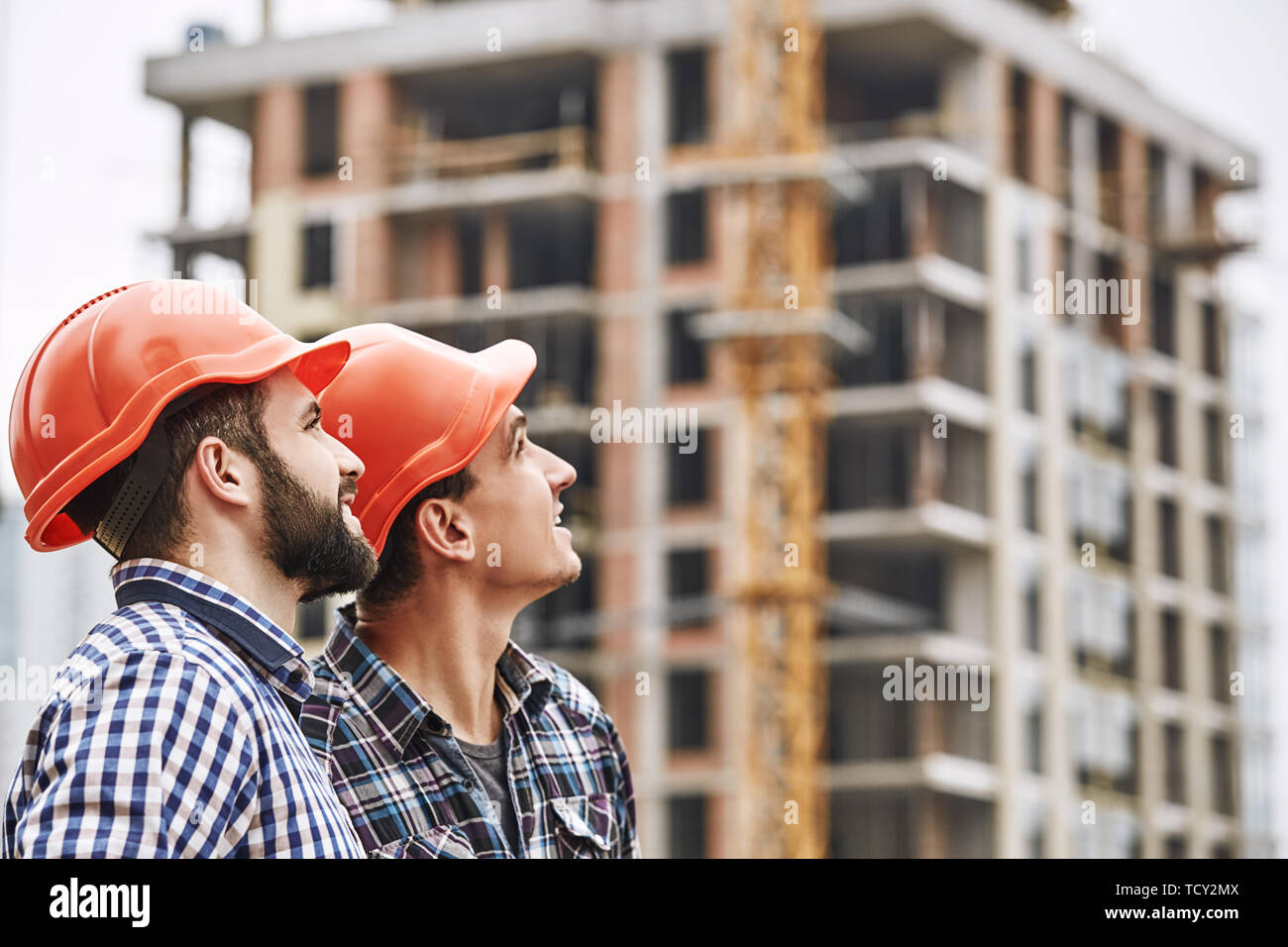 Teamwork. Two young and cheerful builders in red helmets are looking up ...