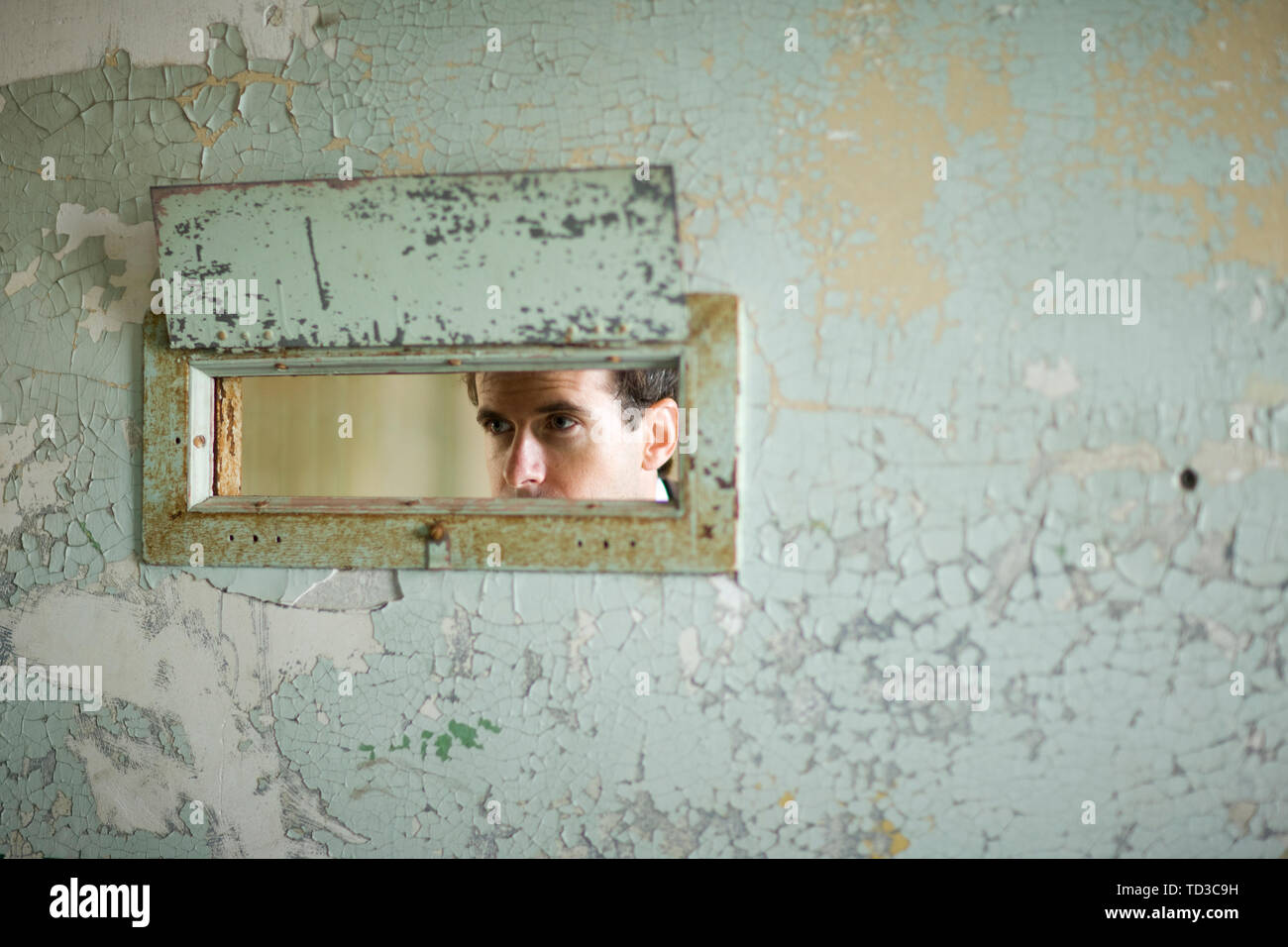 Mid-adult businessman peering through a hole in a wall of a derelict ...