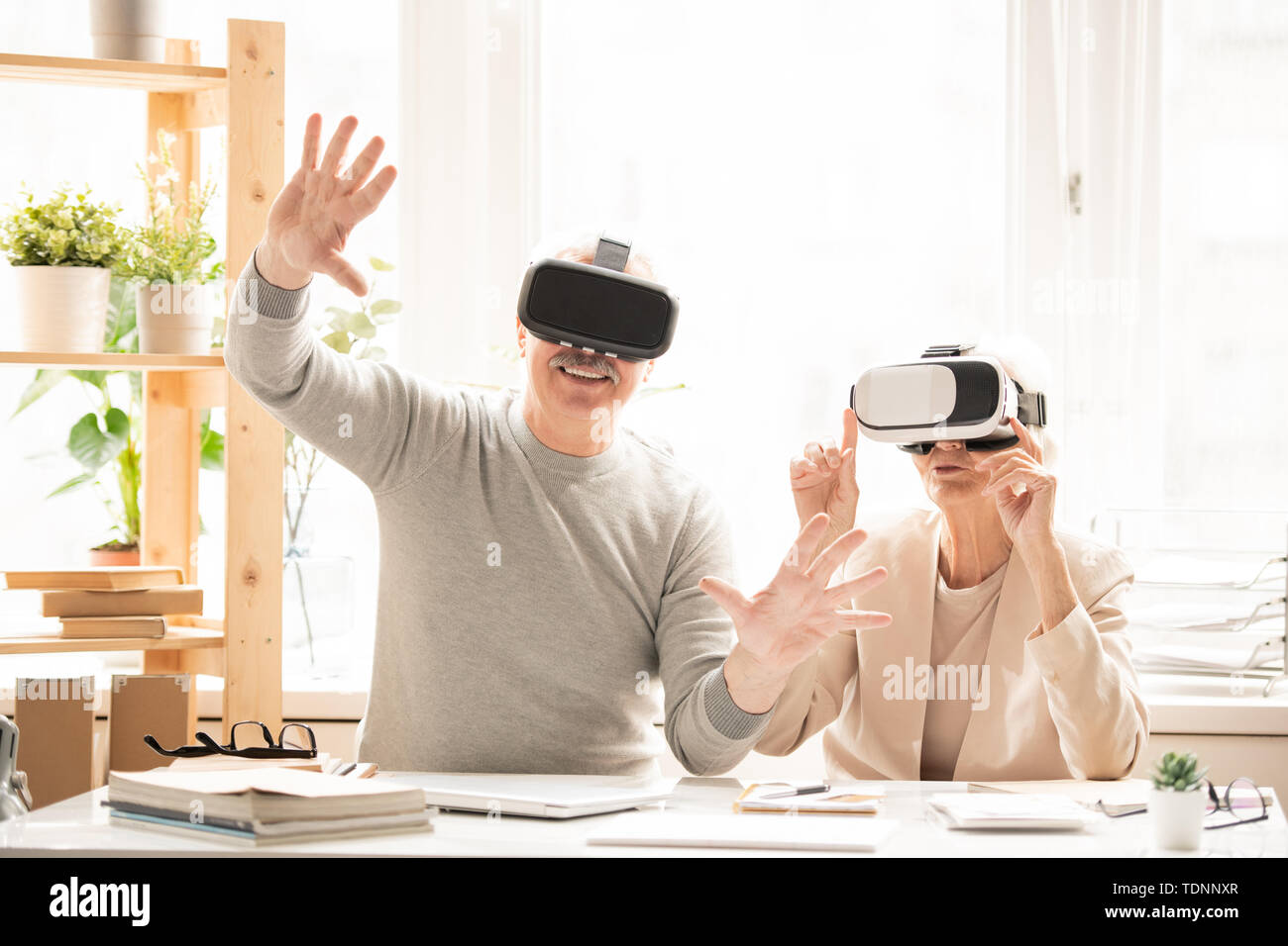 Two contemporary seniors in vr headsets sitting by desk while touching ...