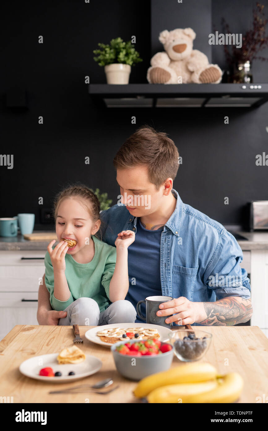 Content hungry little daughter sitting on fathers knees and eating ...