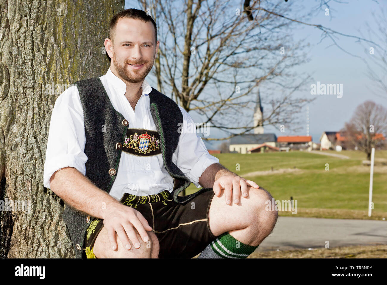 traditional clothed Bavarian man crouching at a tree, church in the ...