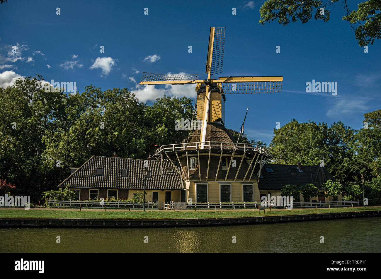House and wooden yellow windmill next to wide tree-lined canal on ...