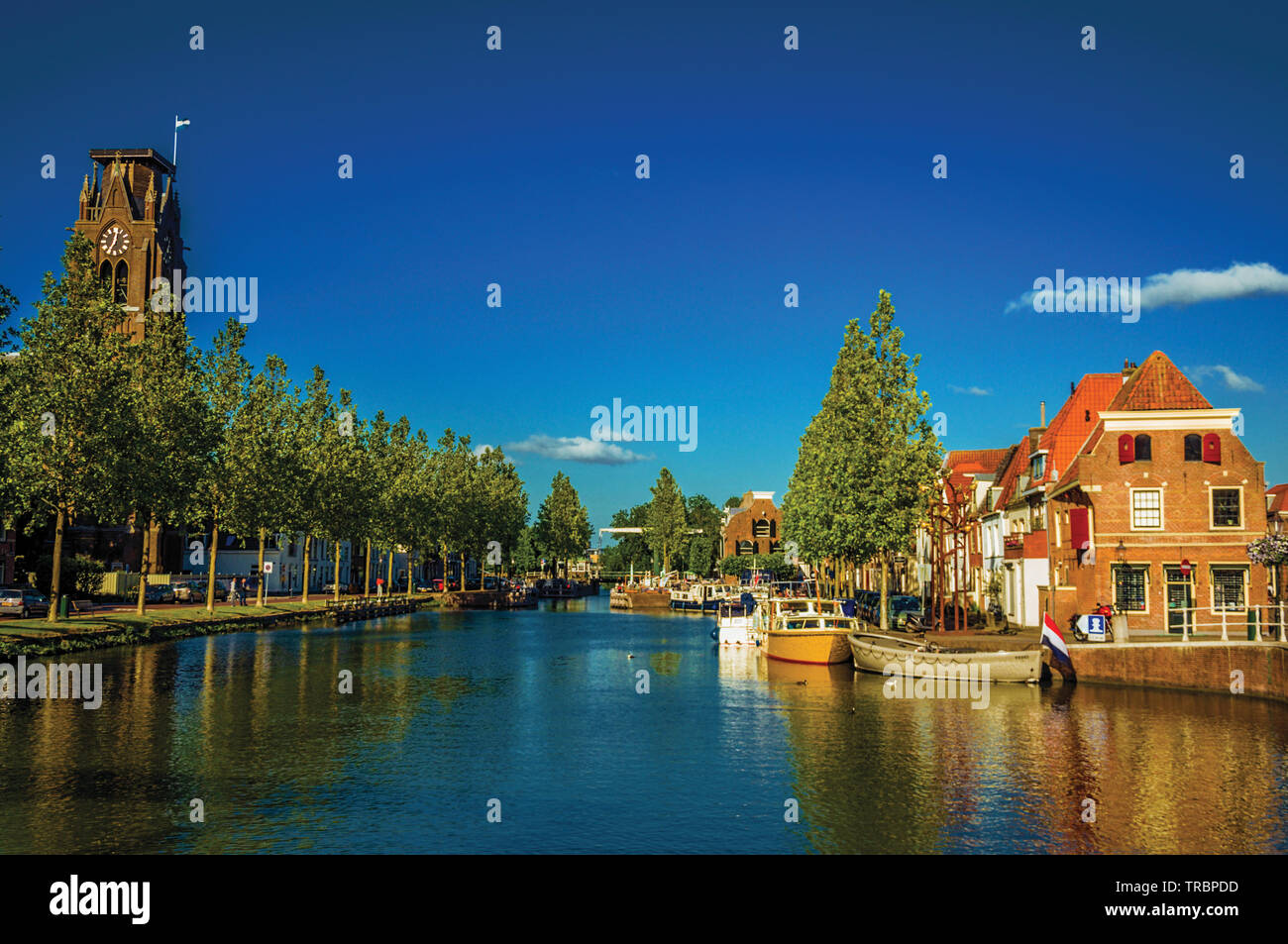 Wide canal with brick houses, boats moored reflected on water at sunset ...