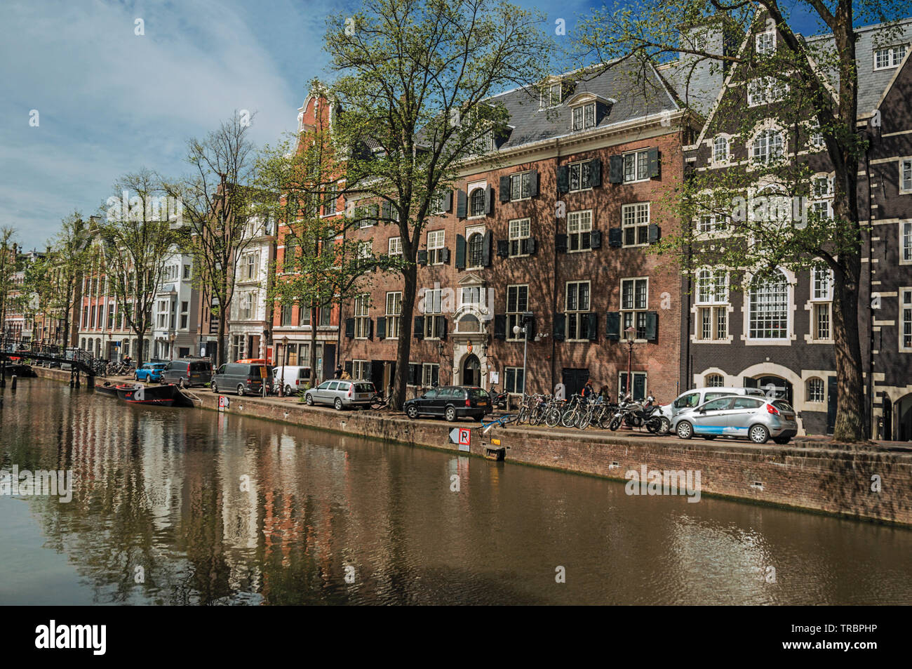 Tree-lined canal with old brick buildings, cars and moored boats in ...