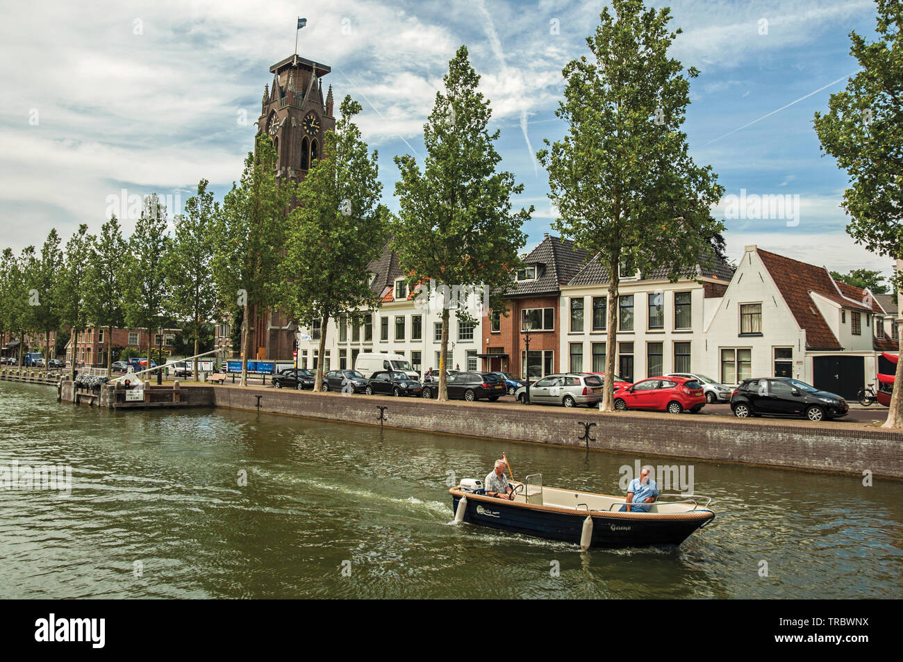 Boat passing by wooded wide canal with brick houses and sunny blue sky ...