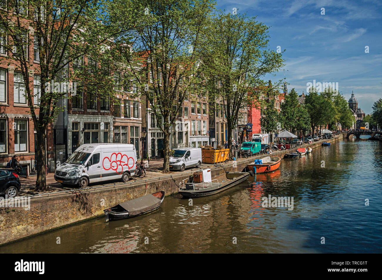Tree-lined canal with old brick buildings, bridge, boats and blue sky ...