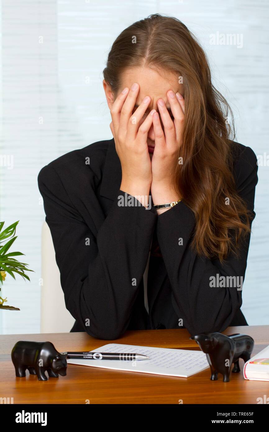 Portrait of a young woman sitting at a desk covering her face with her ...
