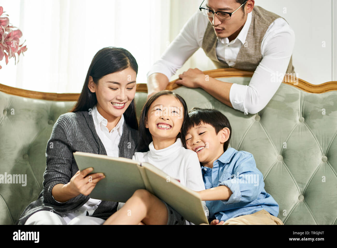 young asian parents and two children sitting on couch reading book together in family living ...