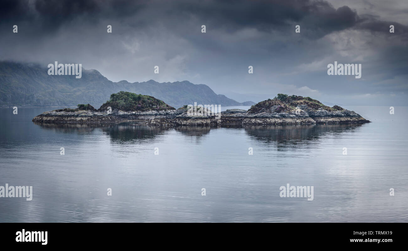 Moody sky over loch with rocky islands and mountain peaks in background ...