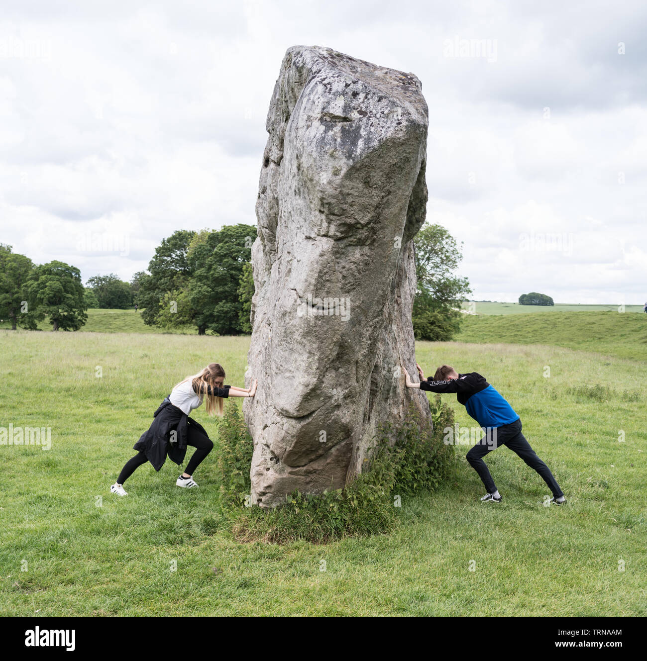 two teenagers pushing against a standing stone at Avebury stone circle ...