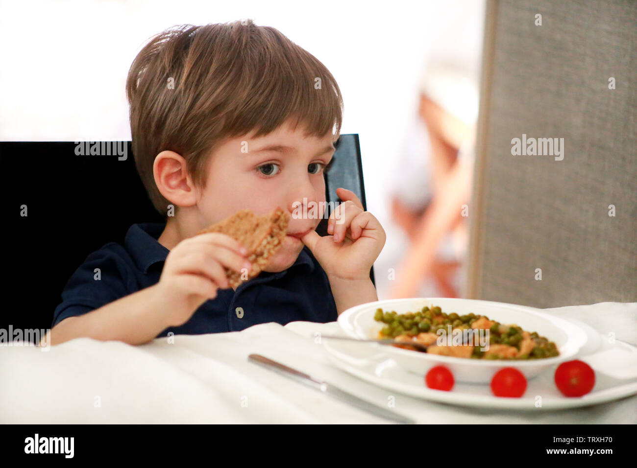 Hungry child sitting in chair at table in kitchen and eating with spoon ...