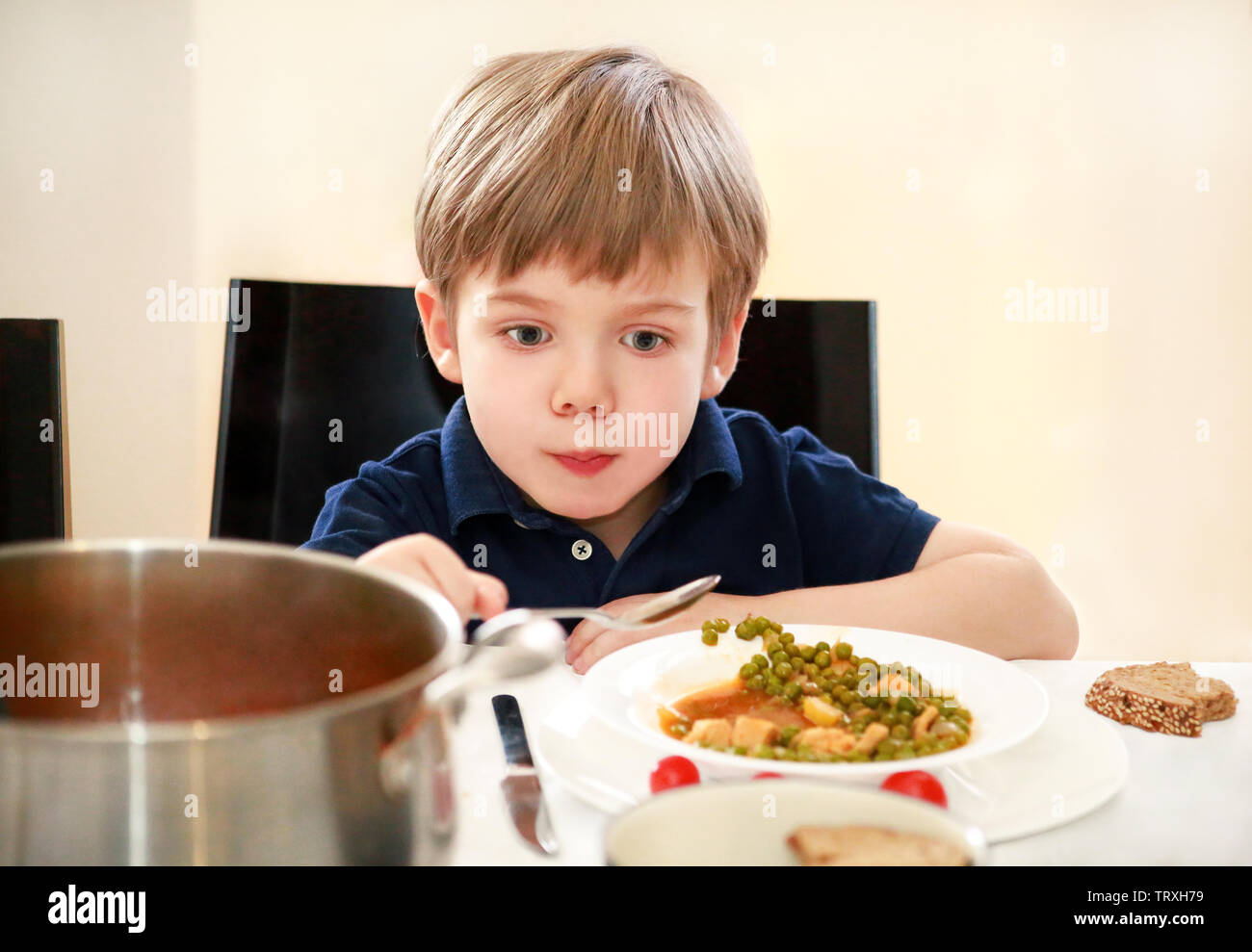 Hungry child sitting in chair at table in kitchen and eating with spoon ...