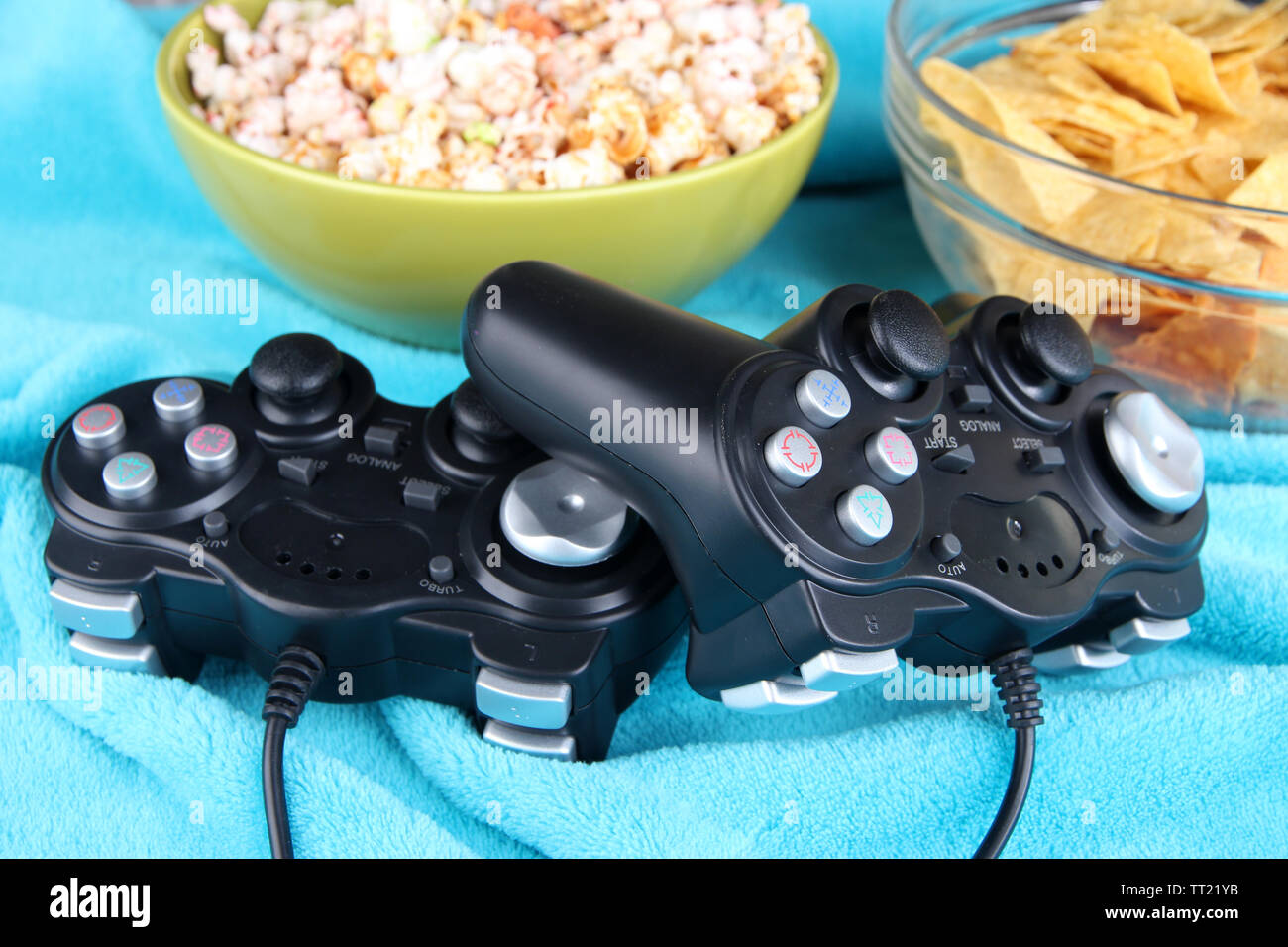 Black game controllers and bowl with snacks on color plaid background ...