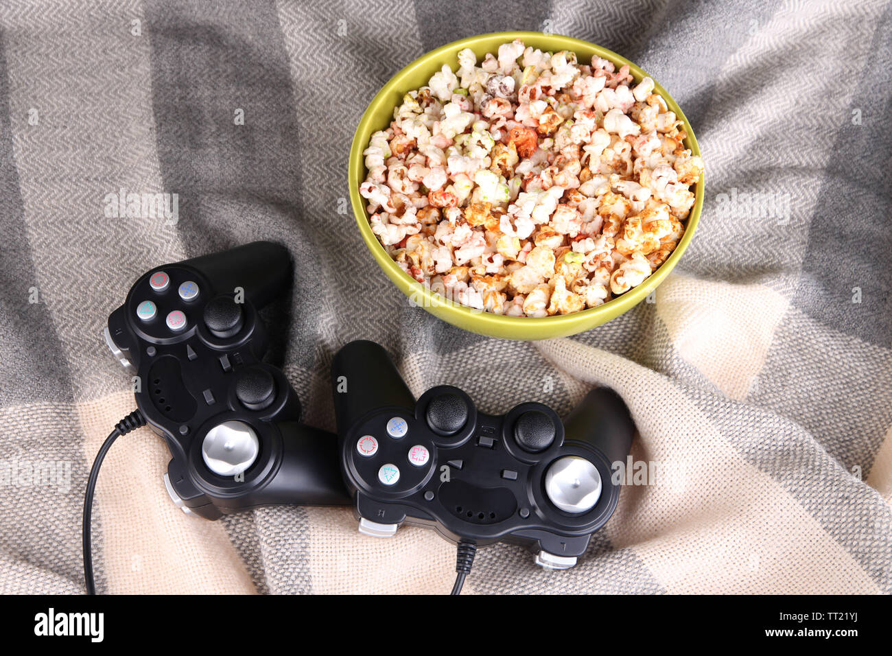 Black game controllers and bowl with snacks on color plaid background ...