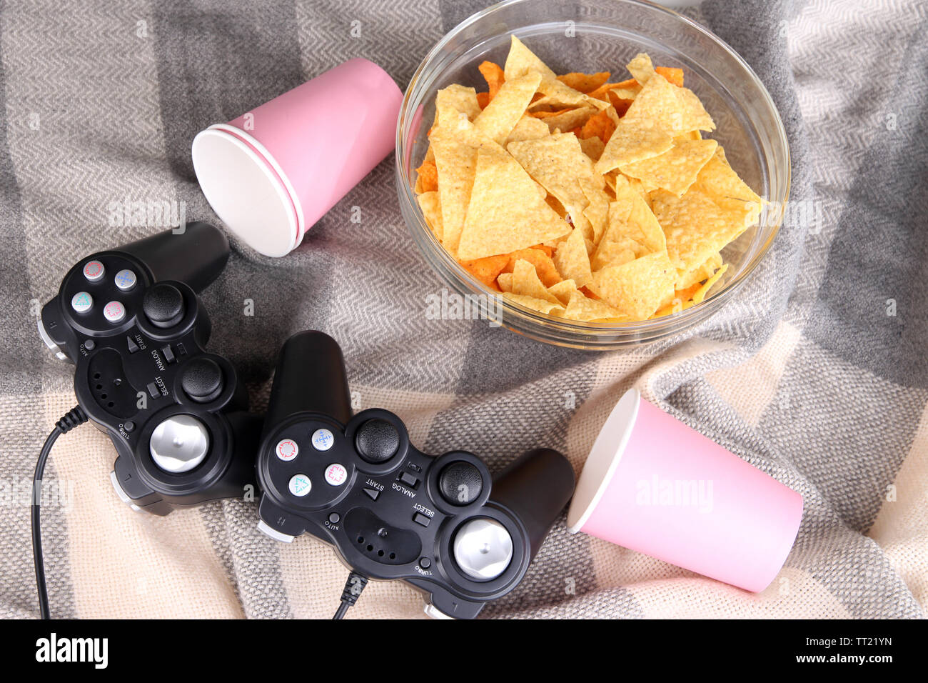 Black game controllers and bowl with snacks on color plaid background ...