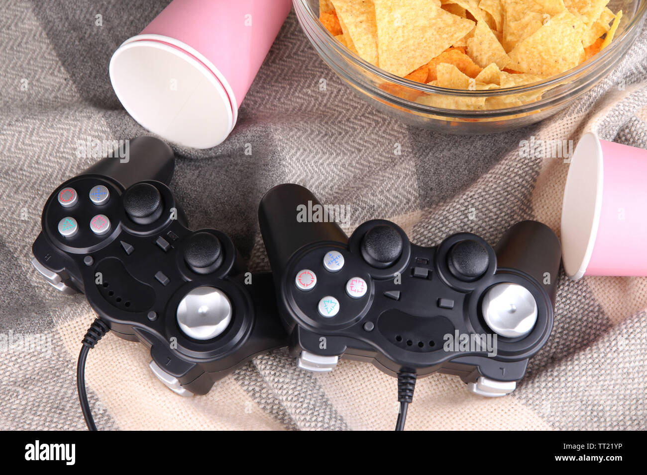 Black game controllers and bowl with snacks on color plaid background ...