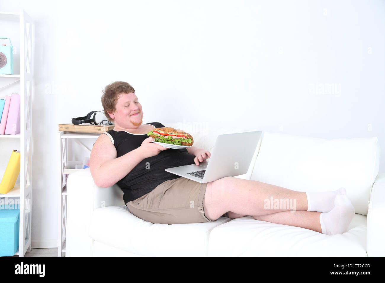 Fat man sitting with laptop on sofa and eating tasty sandwich on home ...
