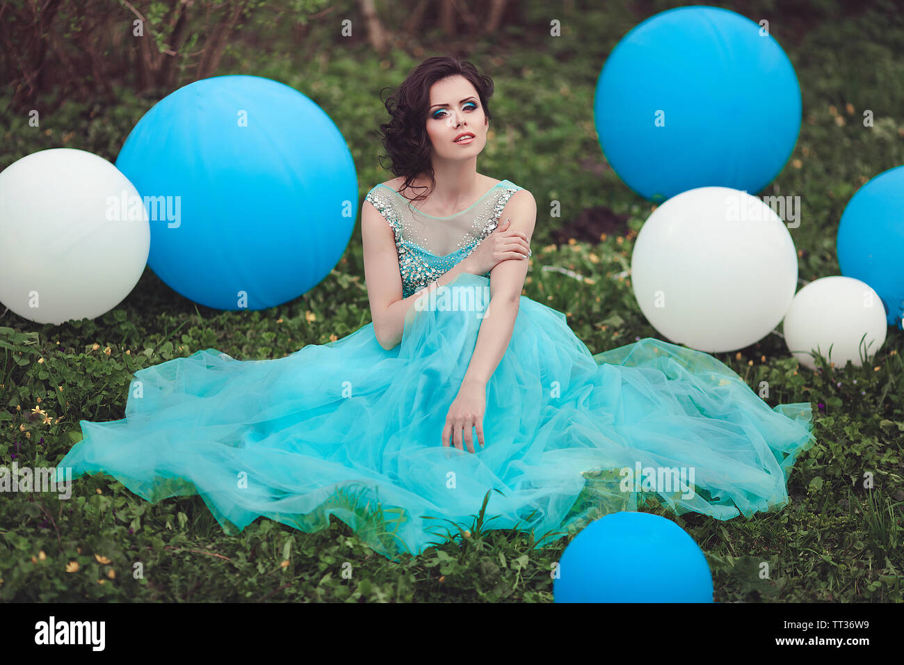 Beautiful girl graduate in a blue dress is sitting on the grass near a ...