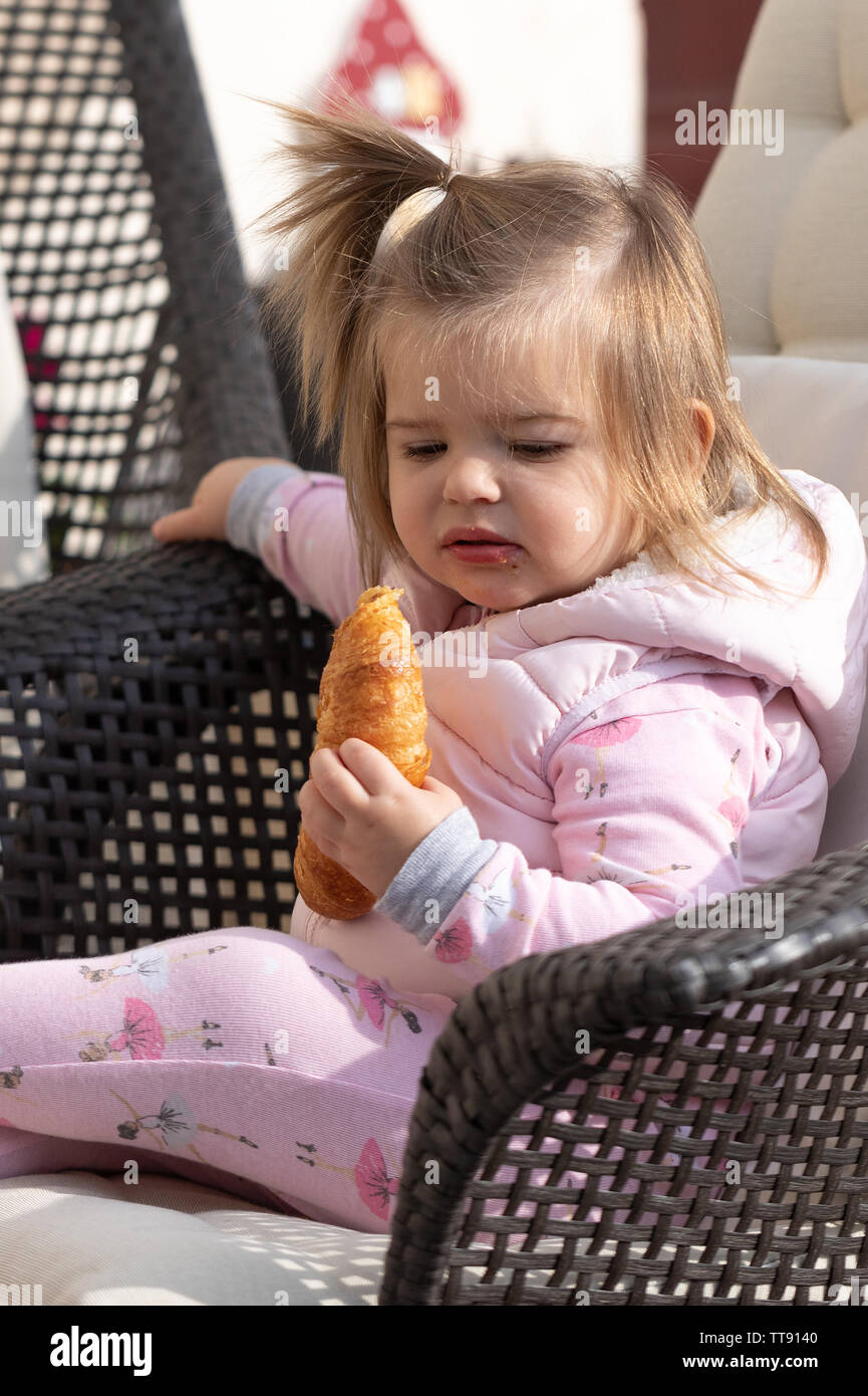 Beautiful baby girl eating the croissant in coffee shop. Breakfast time ...