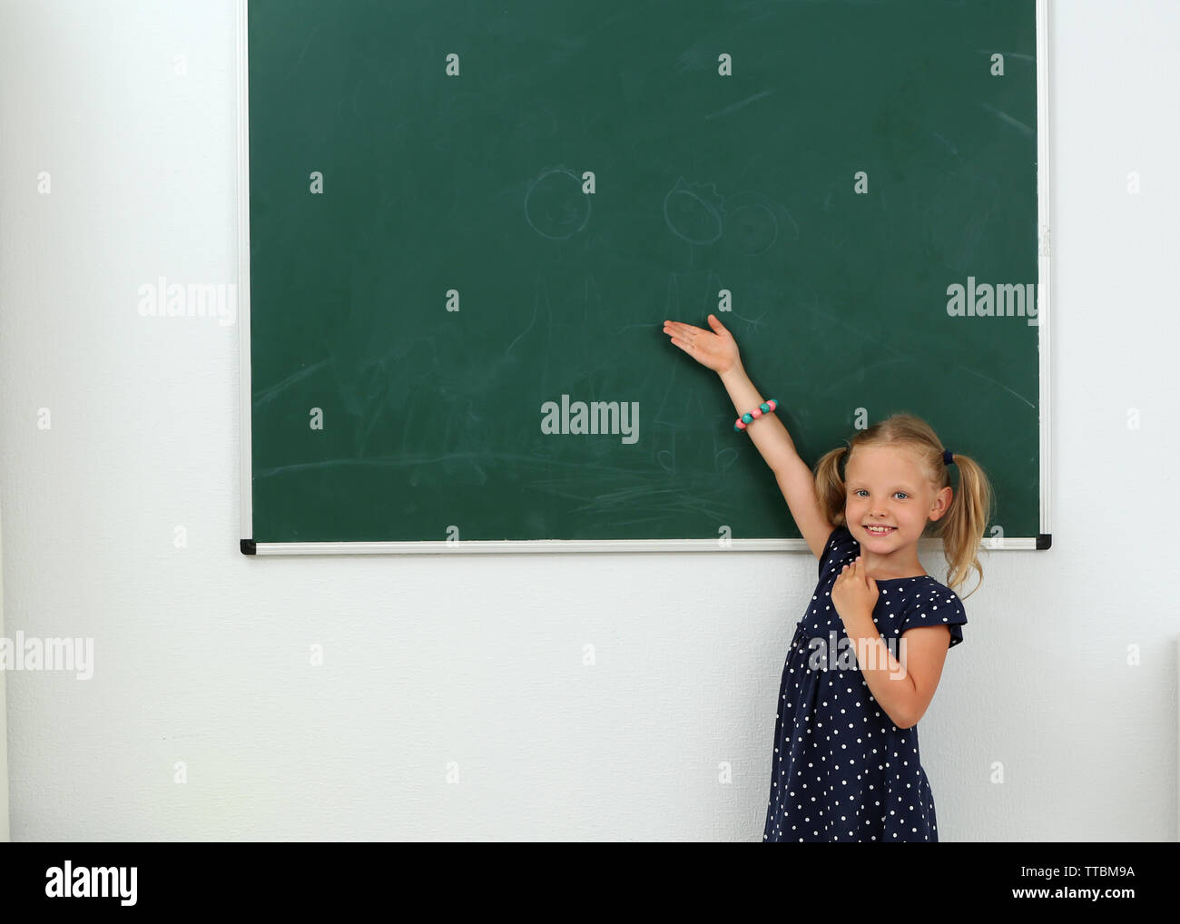 Little girl pointing at something at black chalkboard in classroom ...