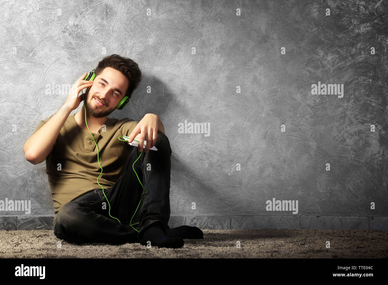 Young bearded man listens music with headphones sitting on grey wall ...