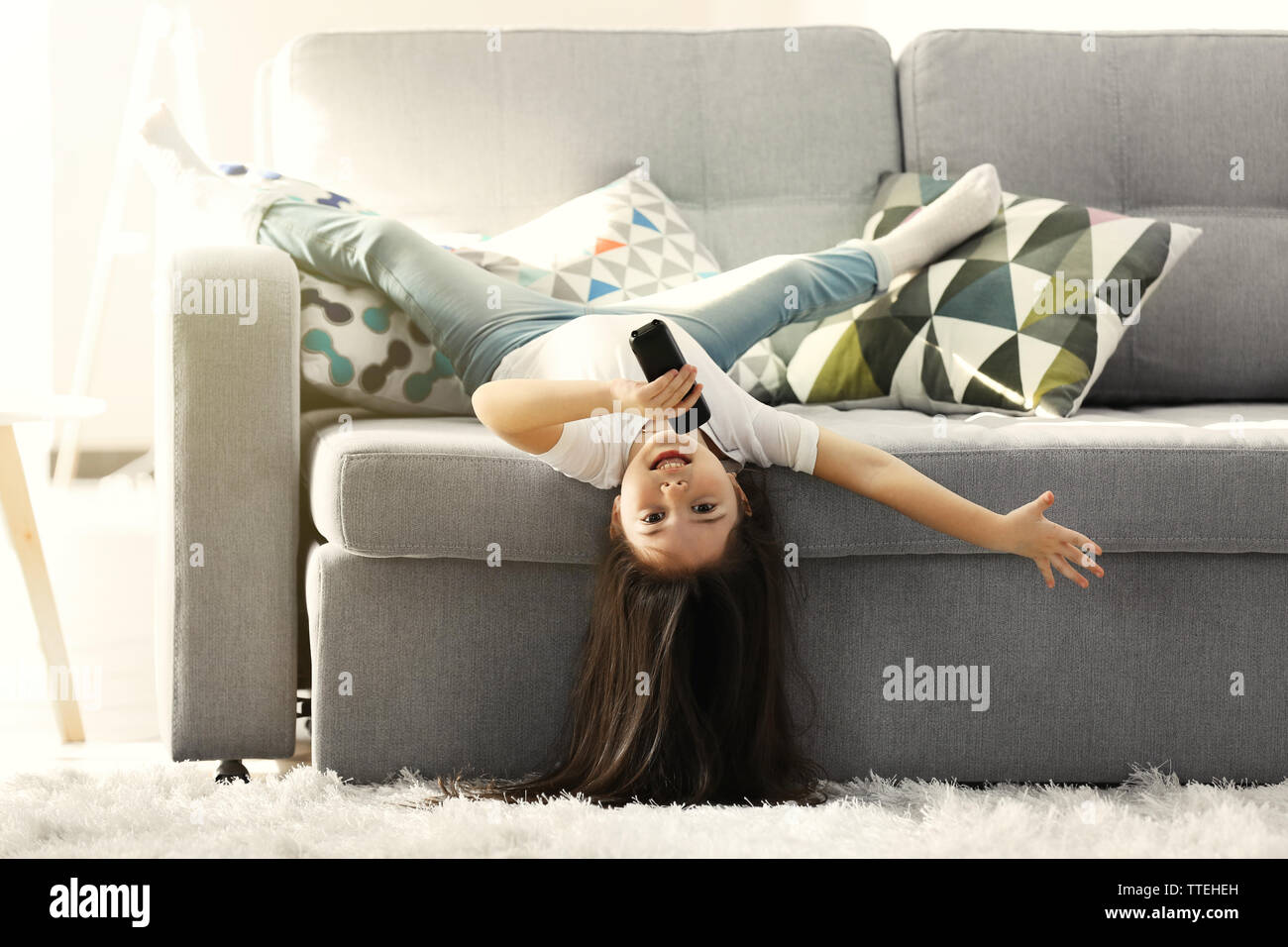 Cute little girl singing in a remote control lying on sofa upside down ...