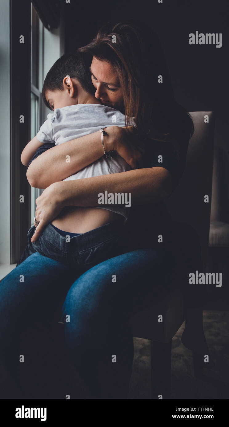 Mother embracing son while sitting on chair at home Stock Photo - Alamy