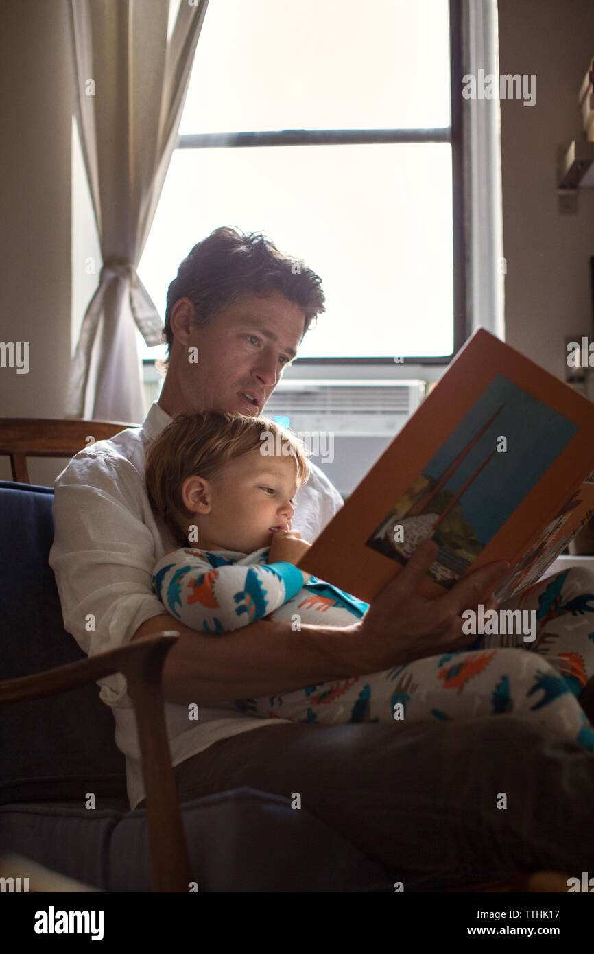 Father reading picture book for son while sitting on chair against ...