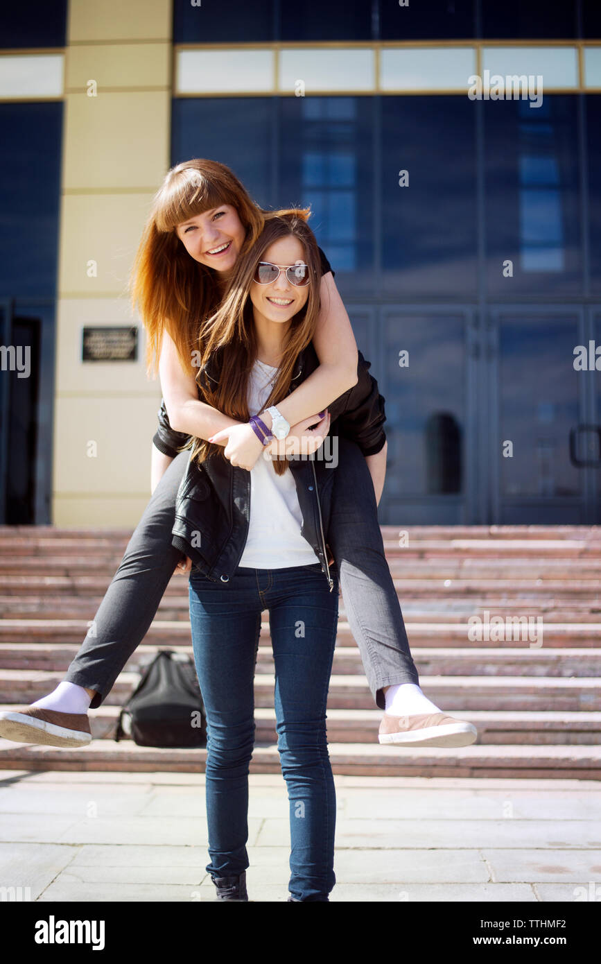 Portrait of cheerful woman giving piggyback to friend outside building ...