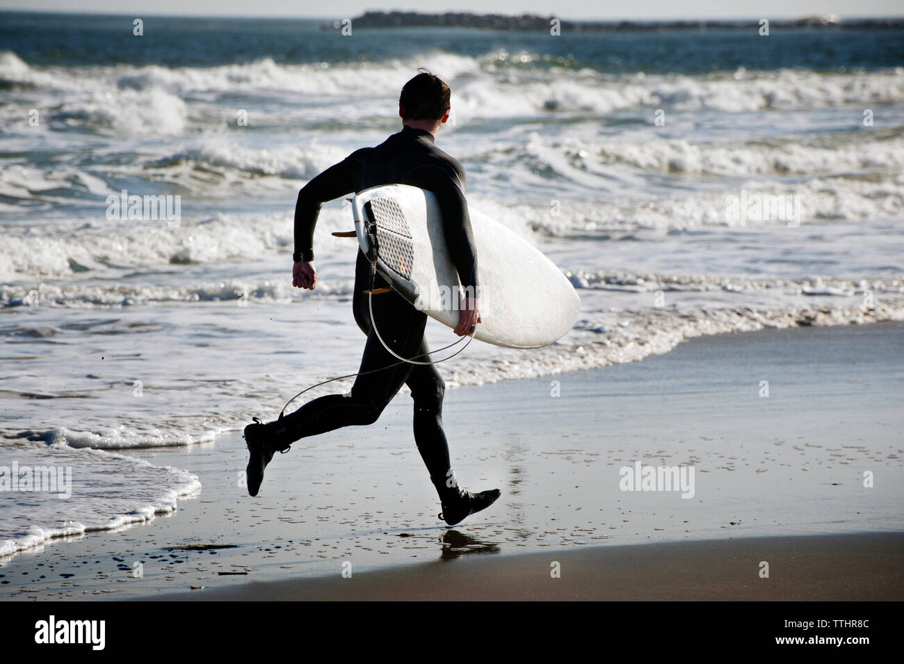 Rear view of surfer carrying surfboard while running on shore at beach ...