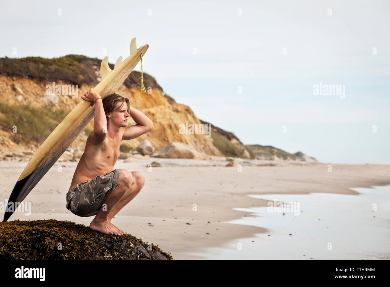 Man carrying surfboard while crouching on rock at beach Stock Photo - Alamy