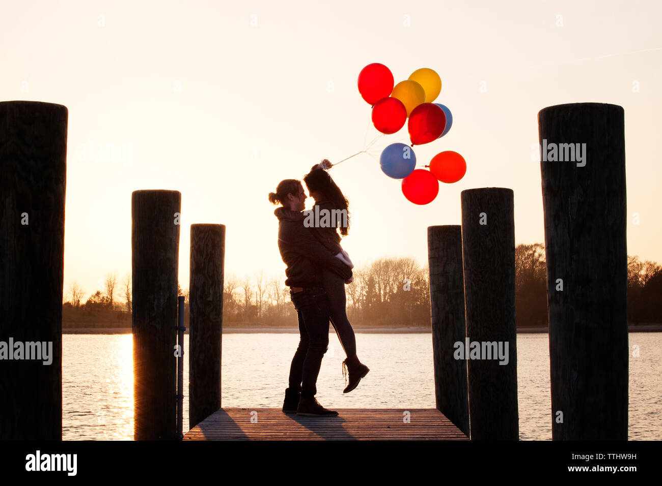 Man lifting woman while holding balloons on pier during sunset Stock ...