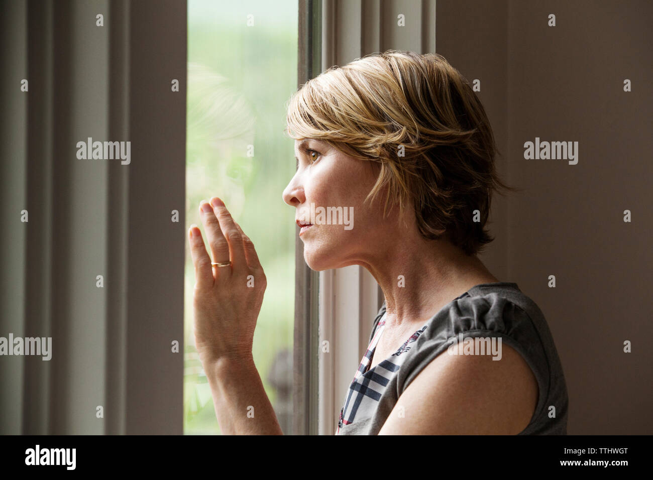 Side view of woman looking through window at home Stock Photo - Alamy