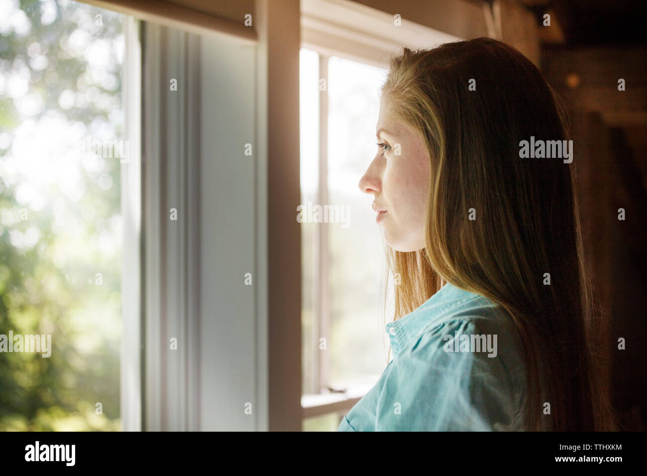 Side view of woman looking through window at home Stock Photo - Alamy